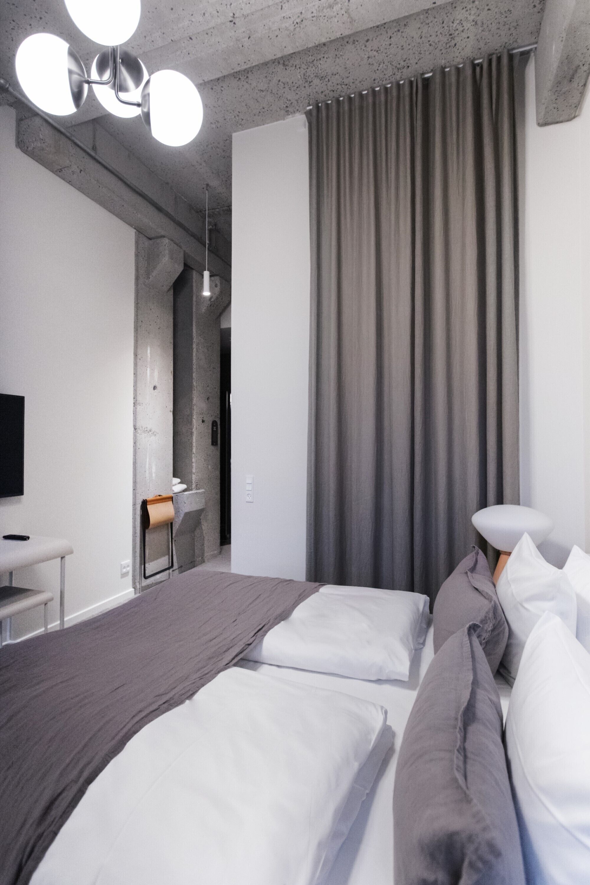 Quarto de hotel moderno com uma cama grande, uma mesinha de cabeceira, uma escrivaninha e um banheiro visível por uma porta aberta.