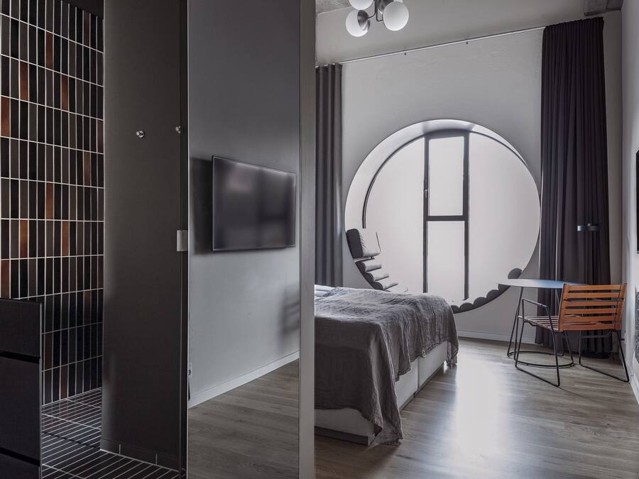 Um quarto moderno com uma cama, uma janela redonda, uma televisão e um espaço de refeições com uma cadeira.