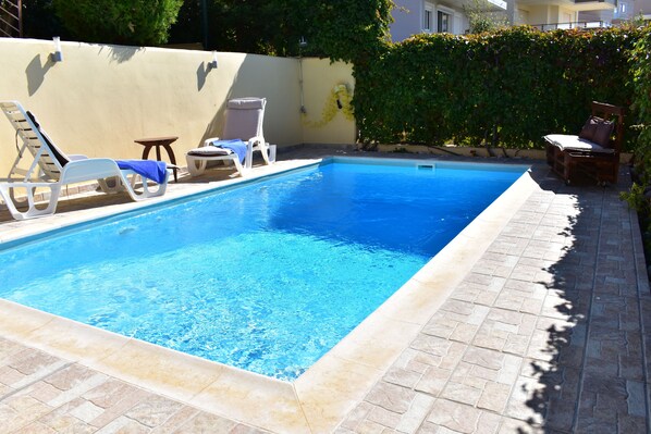 Uma piscina retangular com borda revestida, rodeada por espreguiçadeiras e uma pequena mesa.