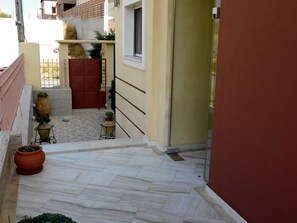 Um corredor estreito ao ar livre, com piso de azulejos, um portão vermelho e plantas em vasos.
