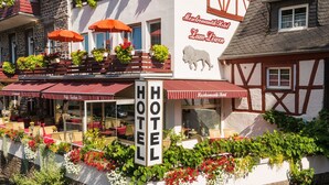 Um hotel tradicional europeu com um letreiro chamativo 'HOTEL', área de estar ao ar livre com guarda-sóis e uma mistura de estilos arquitetônicos em branco e marrom.