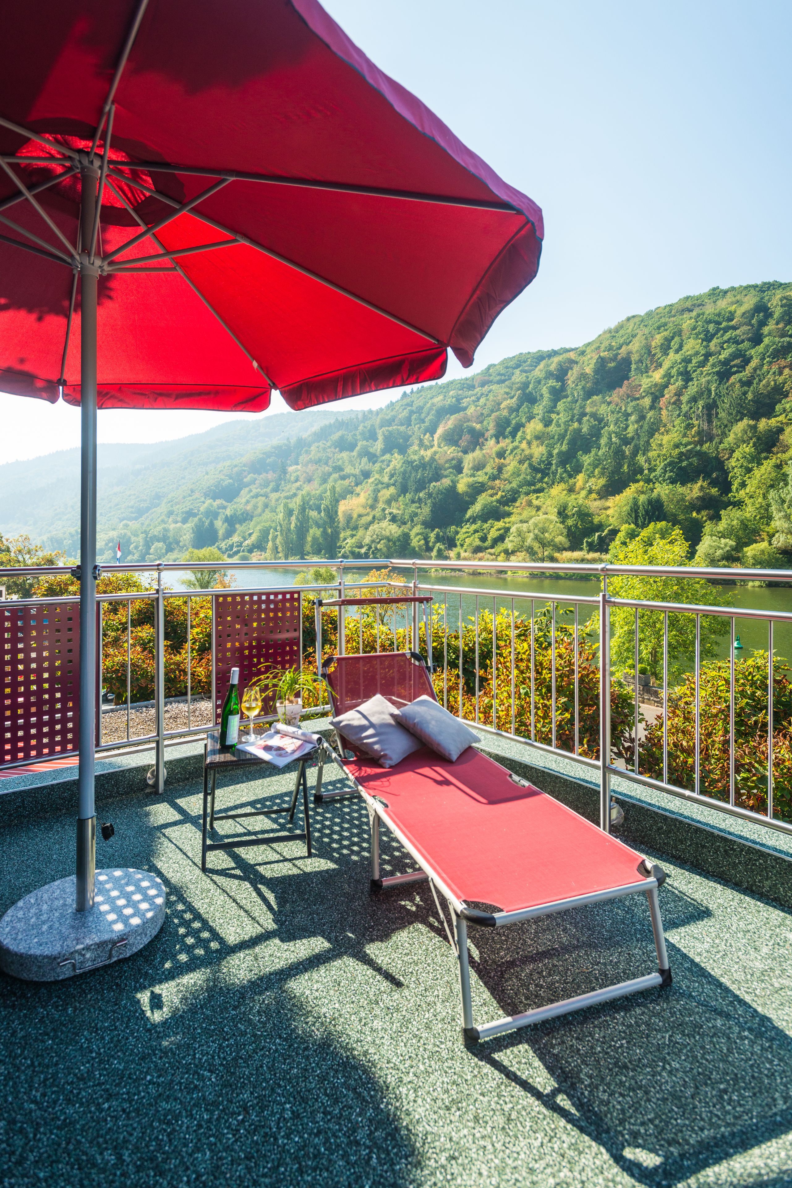 Uma cadeira de descanso vermelha e uma mesa sob um guarda-sol vermelho numa varanda com vista para uma encosta arborizada.