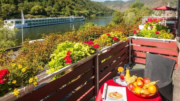 Varanda com cadeiras de vime e uma mesa disposta com frutas e uma bebida, com vista para um rio e colinas.