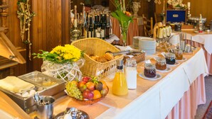 Buffet com diversas opções de comida, incluindo frutas, doces e bebidas, dispostas sobre uma mesa com toalha branca.