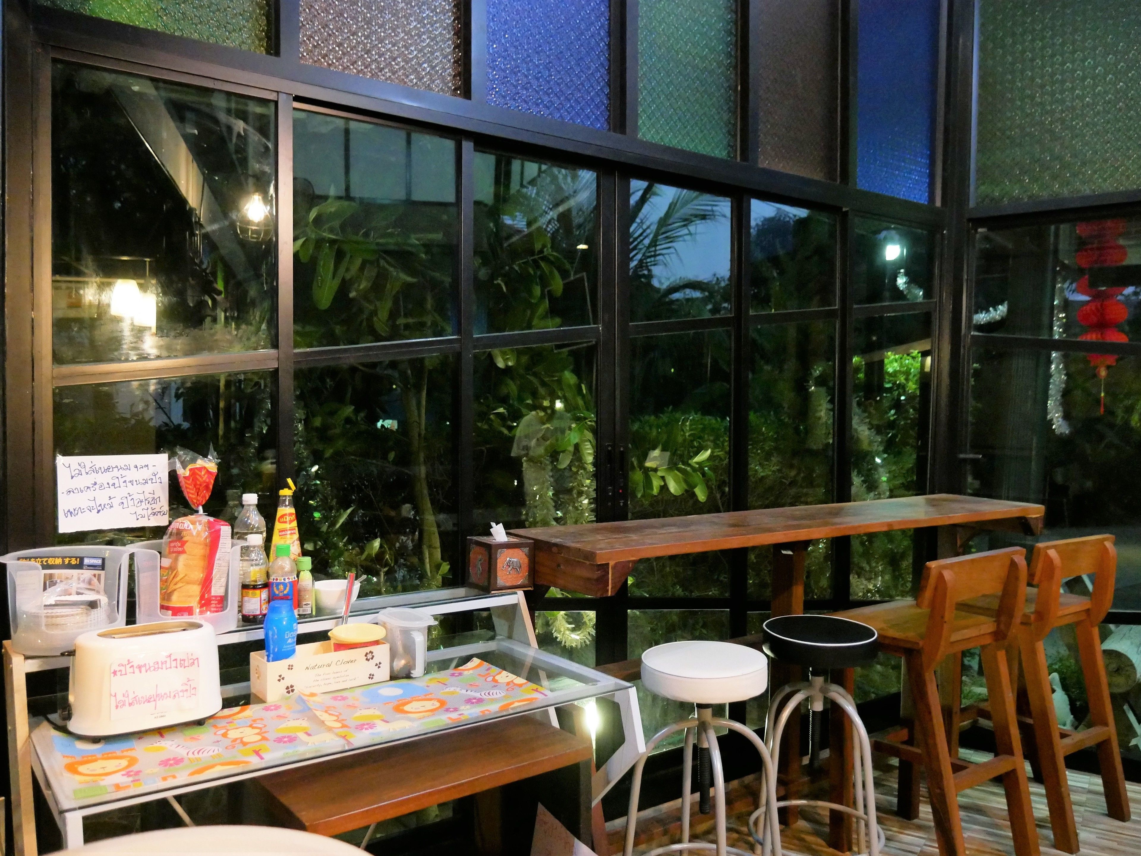 Restaurante com mesa de madeira e banquetas, suporte de menu com itens de comida, e vista para o verde através de grandes janelas.