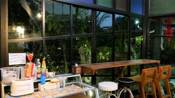 Restaurante com mesa de madeira e banquetas, suporte de menu com itens de comida, e vista para o verde através de grandes janelas.