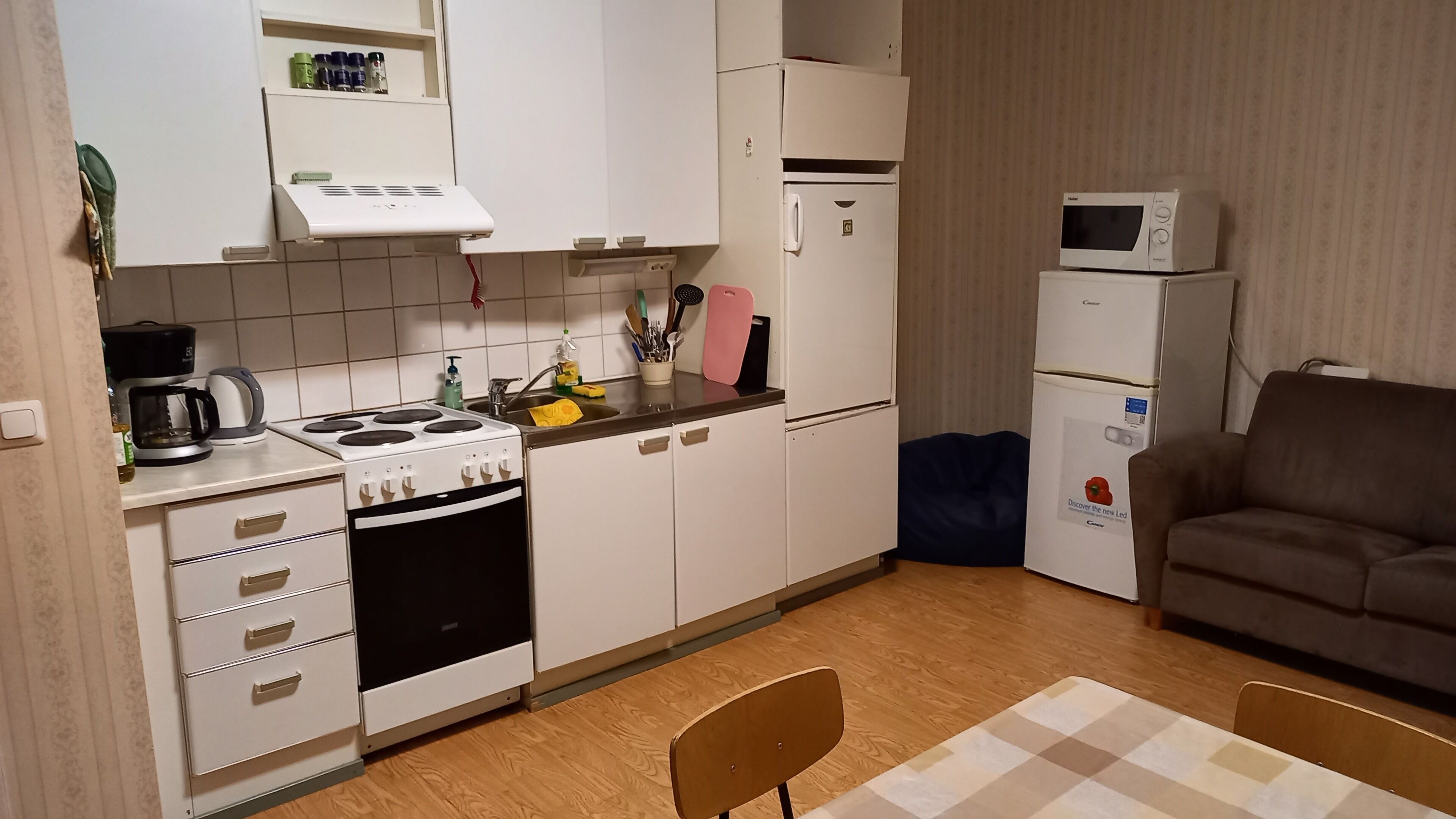 Uma cozinha compacta com fogão, forno, micro-ondas e geladeira. Há uma mesa de jantar com uma cadeira, um sofá e um pequeno pufe azul.