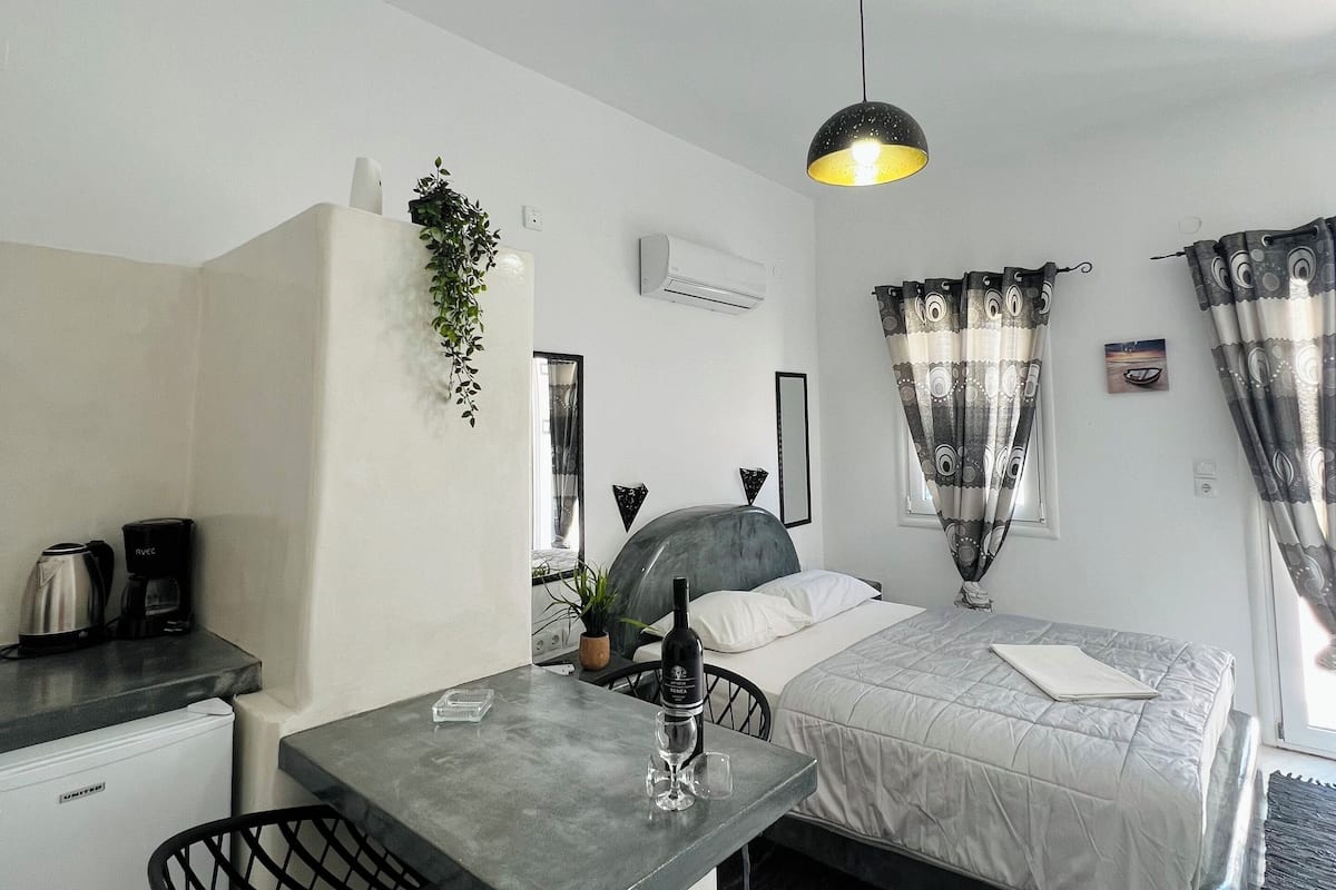 Quarto compacto com cama, mesa de cabeceira, escrivaninha, cadeira e luminária suspensa.