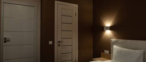 Quarto de hotel com uma cama, mesa de cabeceira, luminária na parede e uma porta.
