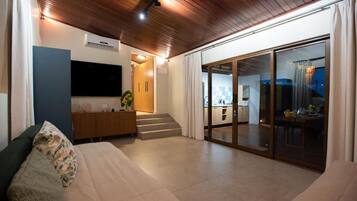 Uma sala de estar moderna com sofá, TV de tela plana, teto de madeira e porta de vidro deslizante que dá para uma cozinha.