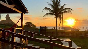 Um deck de madeira com vista para o pôr do sol sobre o oceano, palmeiras e uma montanha ao longe.