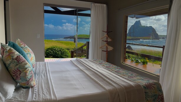 Quarto com cama, vista para o oceano e varanda com grade de madeira.