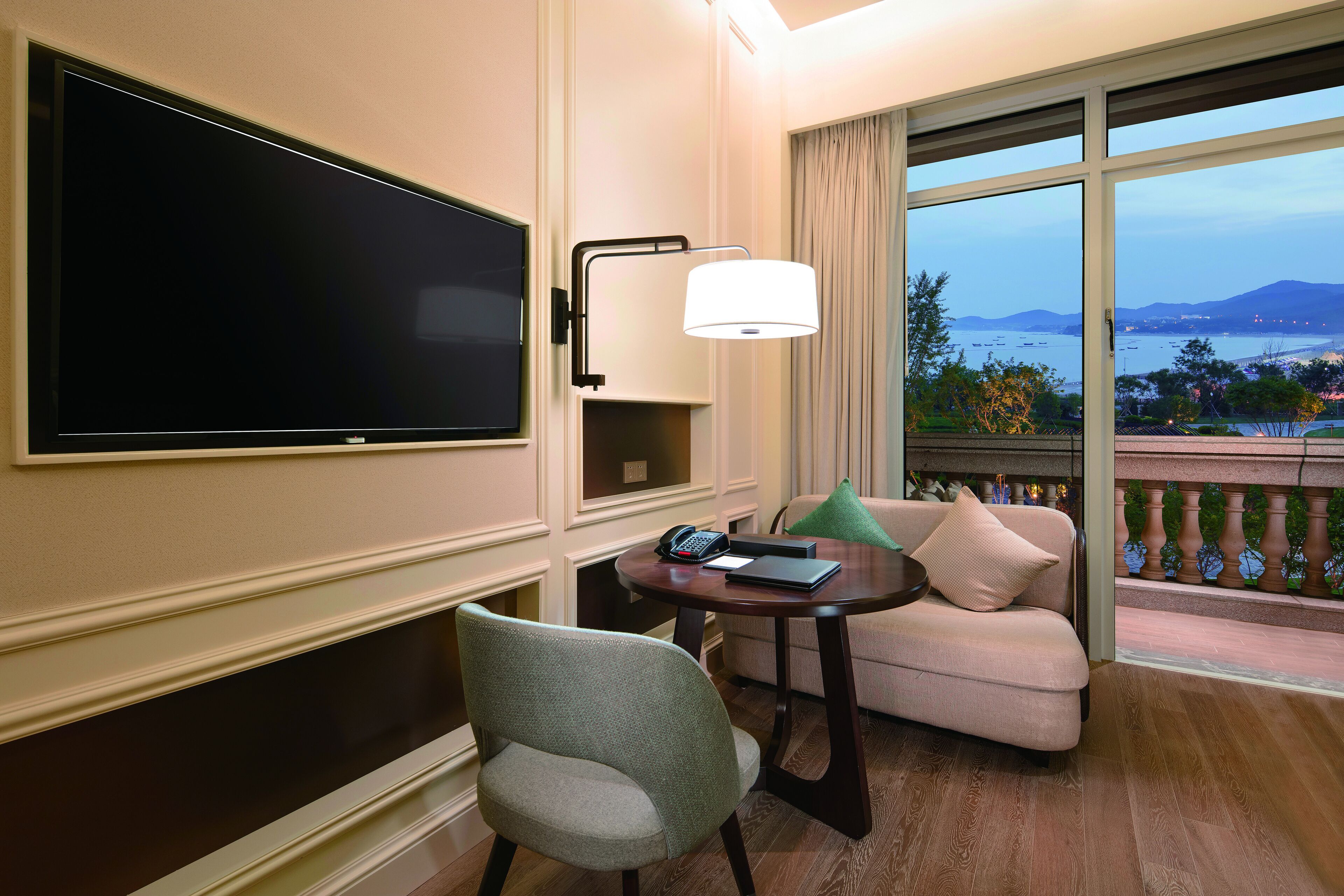 Quarto de hotel com uma televisão de tela grande, uma escrivaninha com luminária, um sofá e uma varanda com vista para o mar e montanhas.