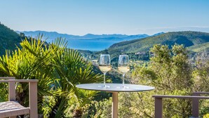 Mesa com duas taças de vinho em terraço com vista para paisagem cênica, com colinas e um corpo de água.
