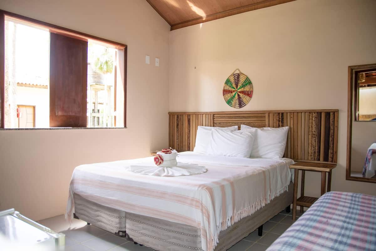 Quarto de hotel com uma cama grande, cabeceira de madeira, mesinha de cabeceira e uma janela com vista, além de um objeto decorativo na parede.