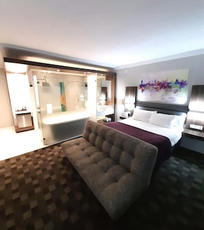 Quarto de hotel com uma cama grande, um sofá, banheiro com chuveiro de vidro e um quadro na parede.