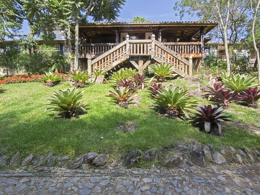 Uma escadaria de madeira leva a um edifício com varanda, rodeado por um jardim com diversas plantas e um caminho de pedra.