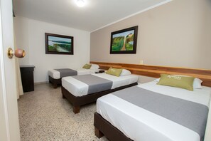 Quarto de hotel com três camas, cada uma com cabeceira de madeira, roupa de cama branca e almofada verde com o nome do hotel. Há duas imagens emolduradas nas paredes, e o chão é coberto por carpete.
