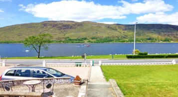 Vista panorâmica de um lago com um barco, um carro e uma área gramada com cerca e um banco.