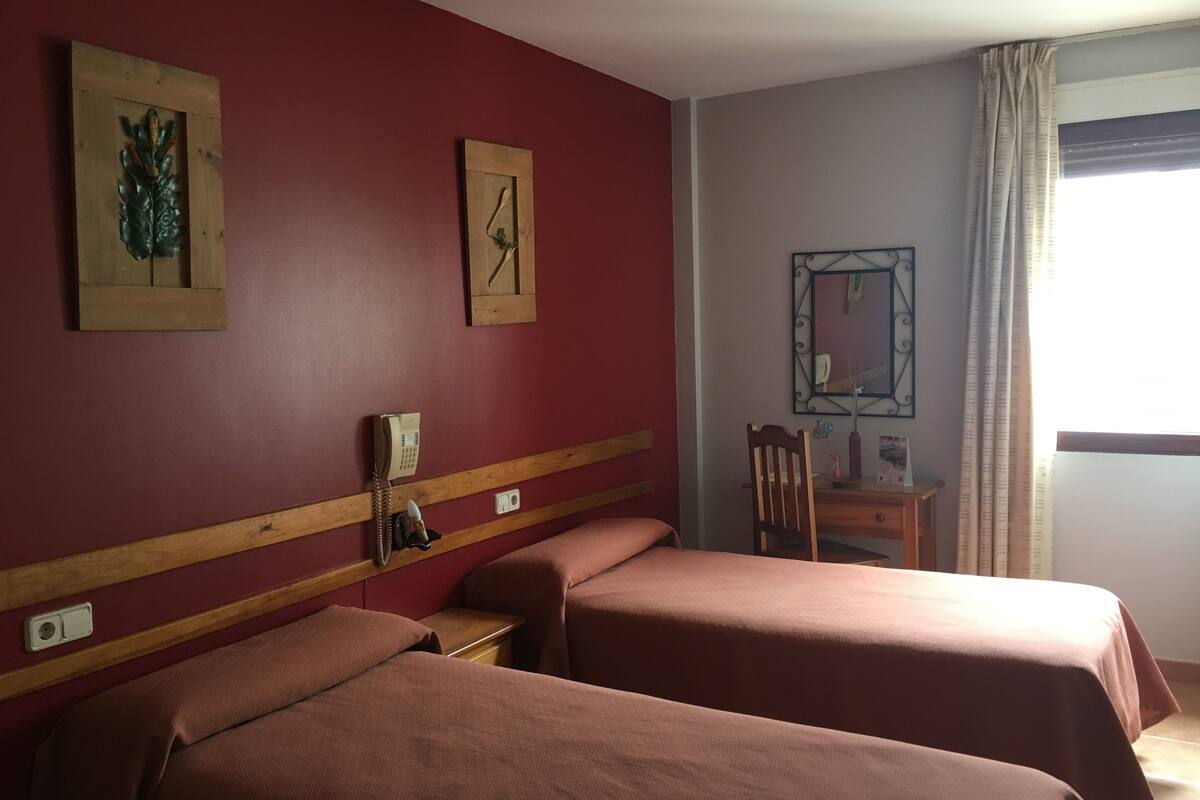 Quarto de hotel com duas camas, uma cabeceira de madeira, uma mesa de trabalho com cadeira, telefone e duas imagens emolduradas na parede.
