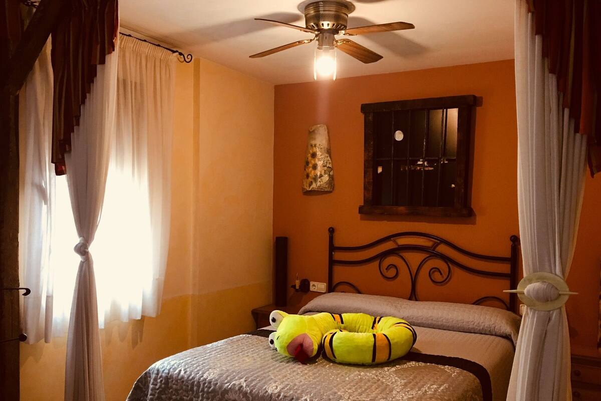 Quarto com cama, ventoinha no teto, janela com cortinas e obra de arte pendurada na parede.
