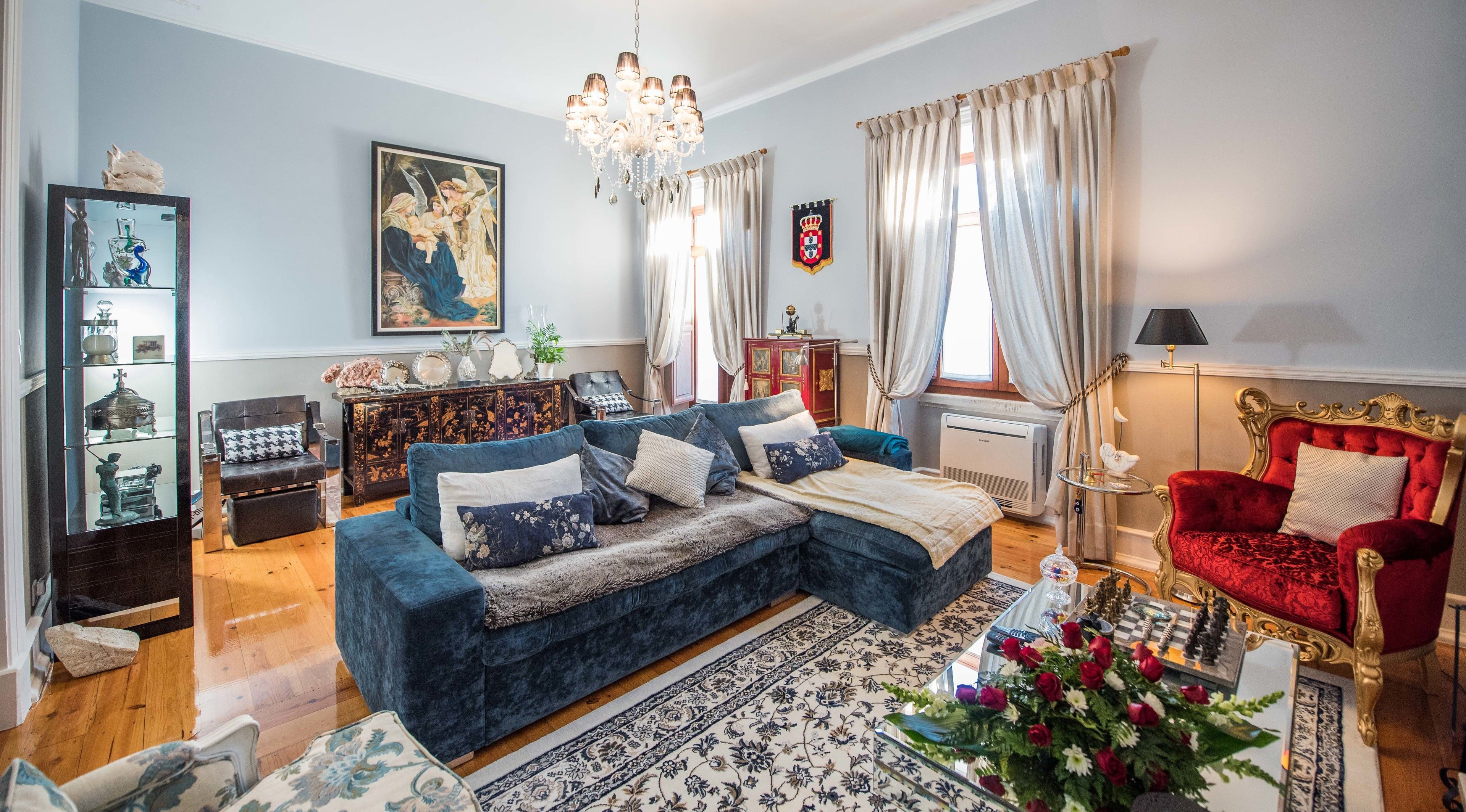 Uma sala de estar com sofá azul, poltrona vermelha, lustre e uma pintura grande.