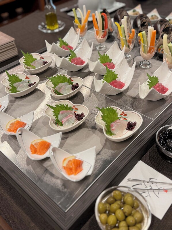 Uma mesa de buffet com diversos pratos, incluindo sashimi e legumes, dispostos sobre uma superfície metálica.