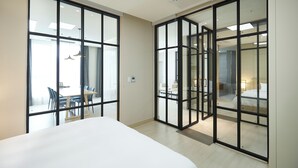 Quarto de hotel moderno com uma cama grande, área de refeições com cadeiras e mesa, e vista através de um painel de vidro para um banheiro e outro quarto.