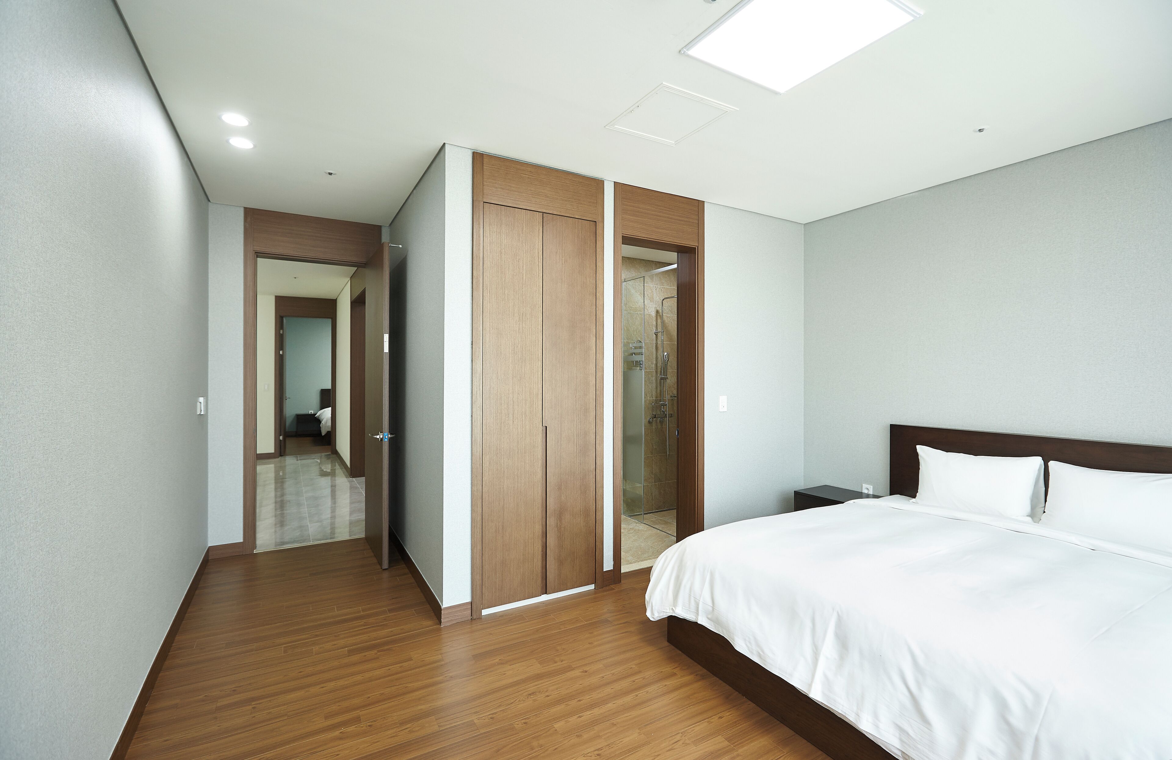 Quarto de hotel moderno com piso de madeira, uma cama grande com roupa de cama branca, um guarda-roupa de madeira e cabine de chuveiro em vidro.