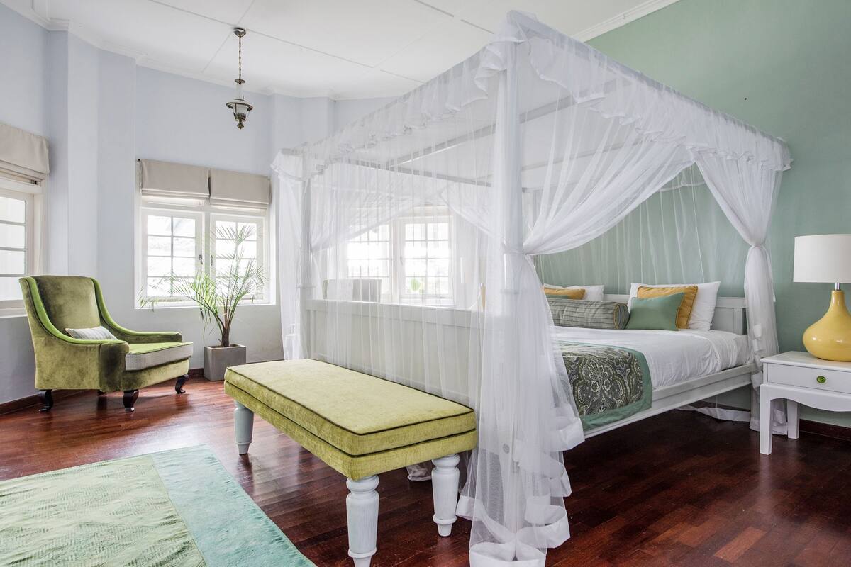 Quarto com cama de dossel, poltrona verde, um banco e mesa de cabeceira com abajur.