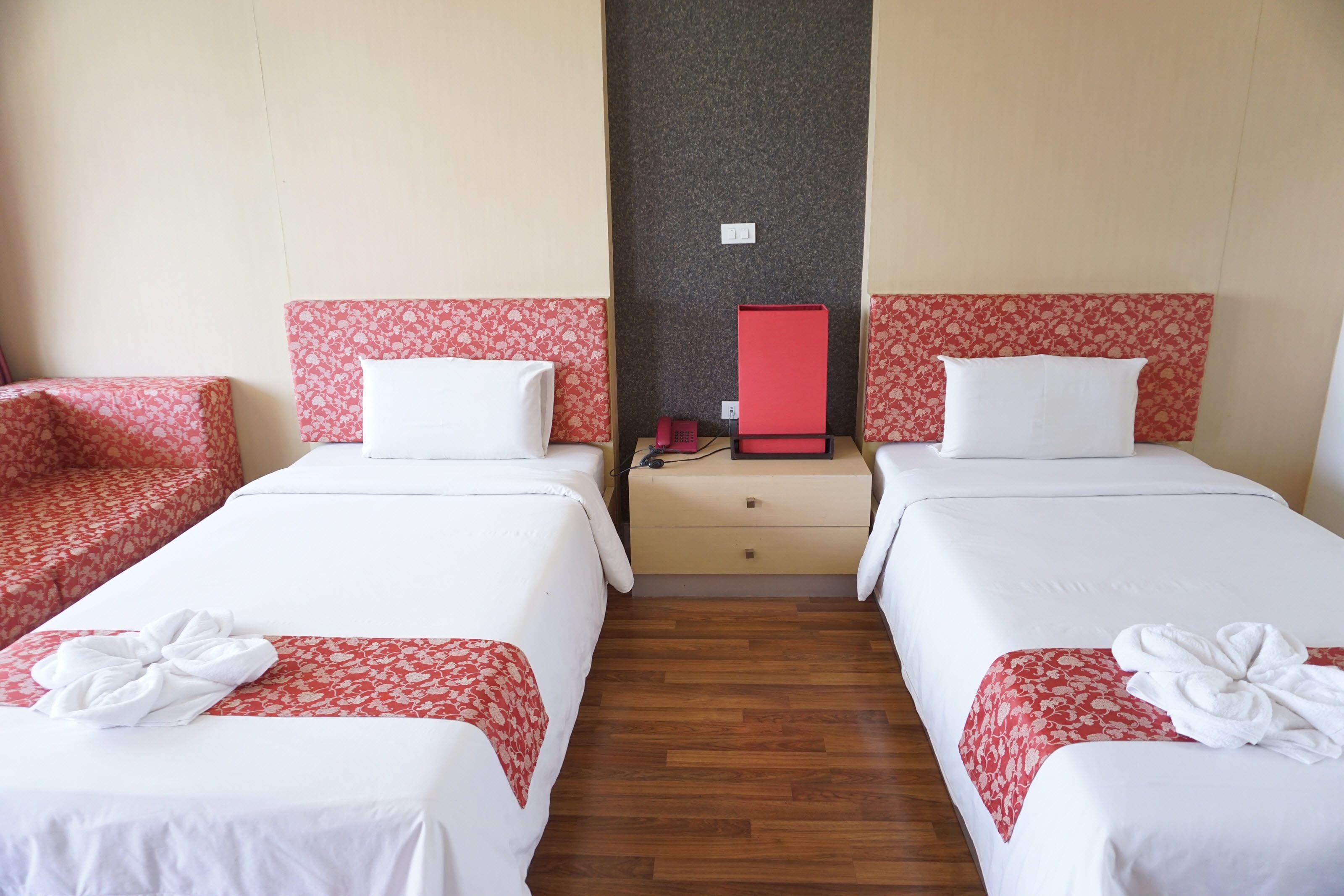 Quarto de hotel com duas camas, cada uma com colcha floral branca e vermelha, piso de madeira e telefone vermelho na mesa de cabeceira.