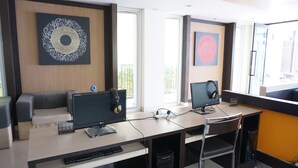 Espaço de escritório moderno com duas mesas, cada uma equipada com um computador, auscultadores e um telefone. Há duas cadeiras, um cesto de lixo pequeno e um teclado nas mesas. O quarto tem uma janela grande, uma obra de arte na parede e um candeeiro no teto.
