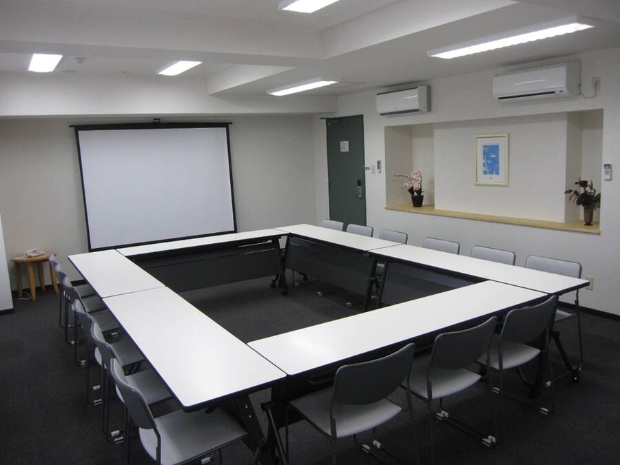 Uma sala de conferências com uma mesa comprida, cadeiras, tela de projetor, aparelho de ar condicionado e uma pequena mesa com um telefone.