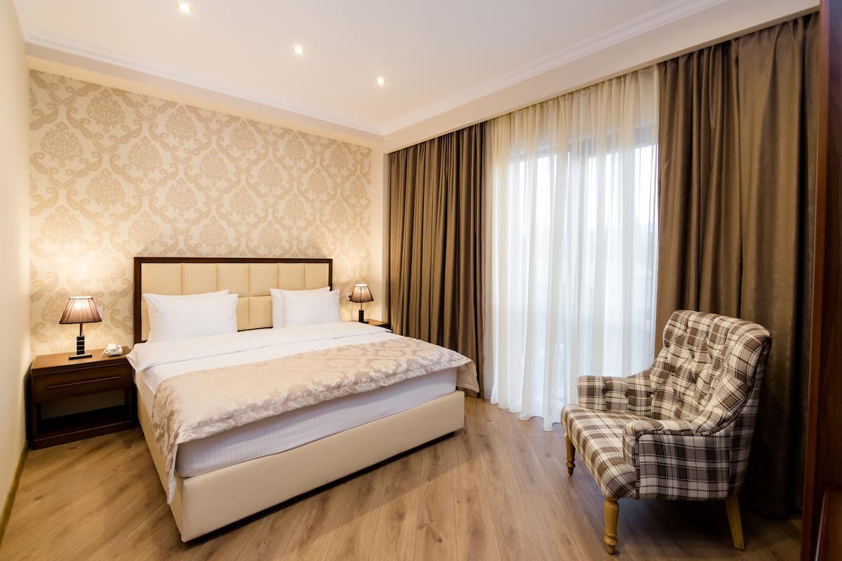 Quarto de hotel com uma cama grande, mesa de cabeceira, uma cadeira e uma janela com cortinas.