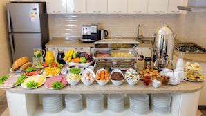 Disposição de buffet com diversos alimentos e bebidas, incluindo frutas, queijos e carnes, dispostos em um balcão com pratos empilhados embaixo.