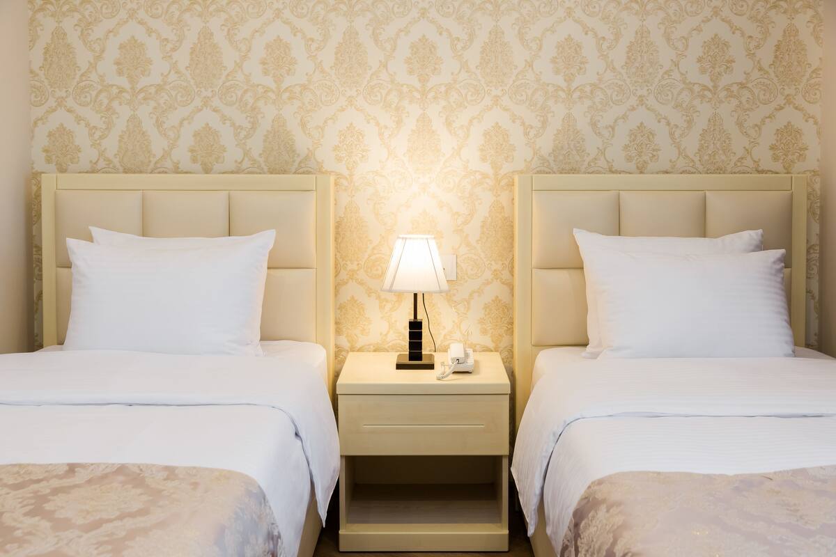 Quarto de hotel com duas camas, uma mesa de cabeceira com abajur e papel de parede estampado.