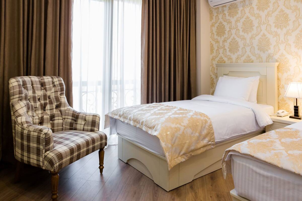 Quarto de hotel com cama, uma cadeira, mesa de cabeceira com abajur e papel de parede estampado.