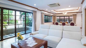 Ampla sala de estar com um sofá branco grande, uma mesa de centro de madeira com um vaso de flores amarelas e uma mesa de jantar posta para seis com cadeiras vermelhas.