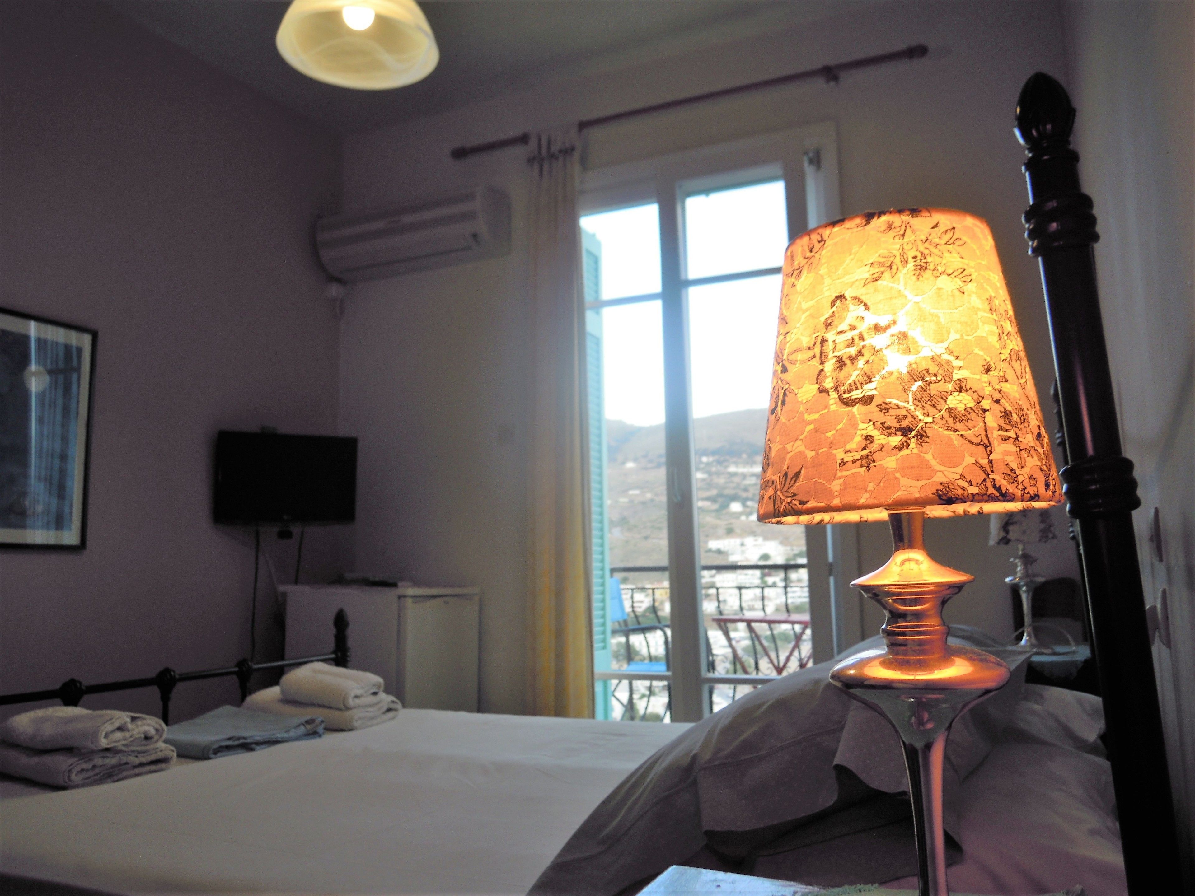 Quarto de hotel com cama, luminária, TV e janela com vista para o exterior.