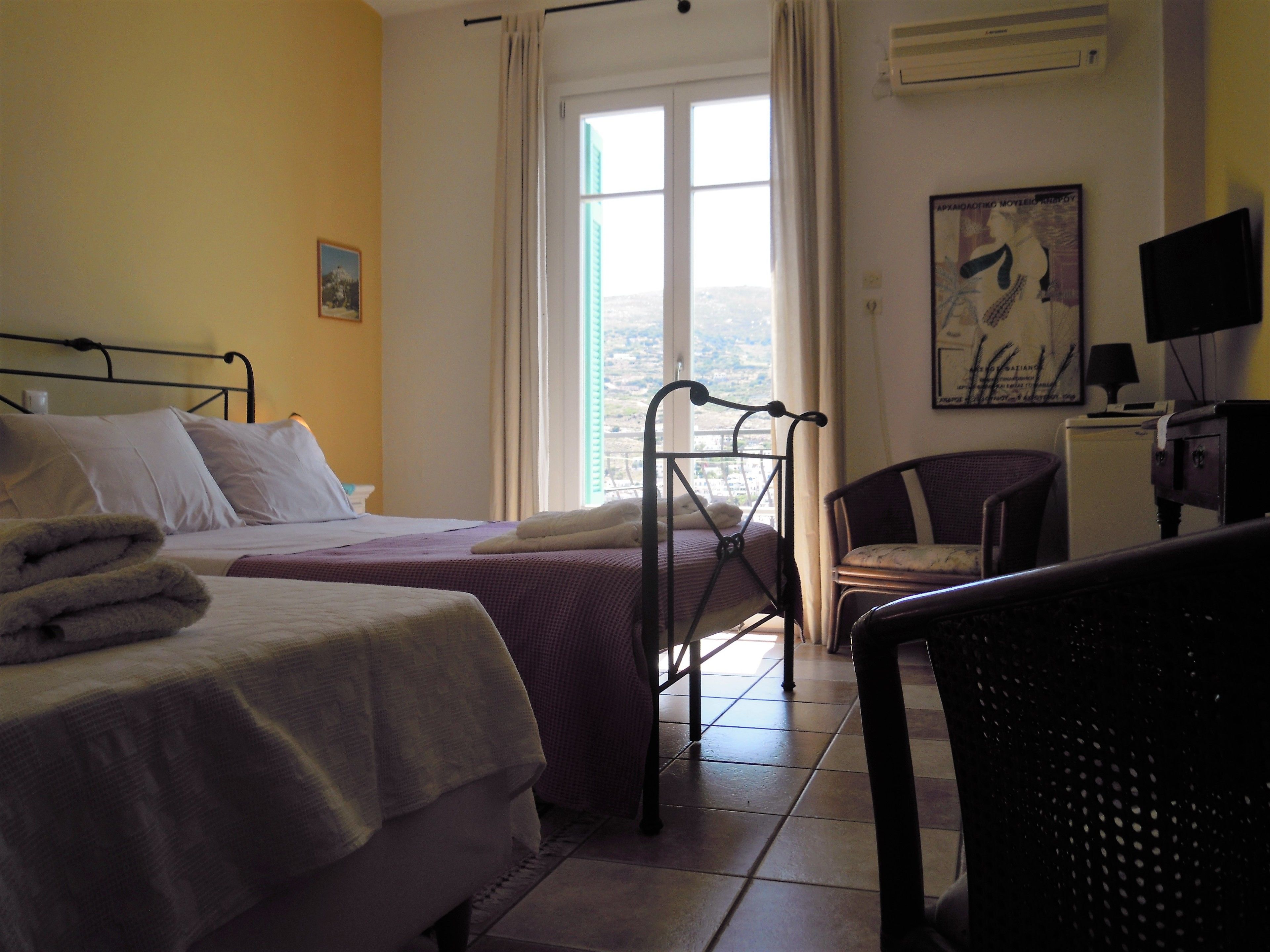 Quarto de hotel com duas camas, uma cadeira, uma escrivaninha com computador e uma janela com vista.