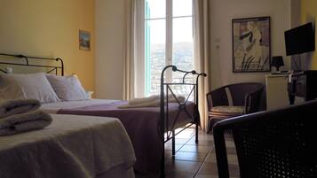 Quarto de hotel com duas camas, uma cadeira, uma escrivaninha com computador e uma janela com vista.