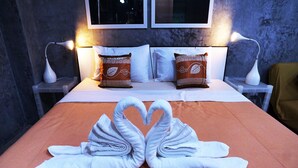 Cama bem arrumada com lençóis brancos e duas almofadas decorativas, mesa de cabeceira com abajur e um porta-retrato fixado na parede.
