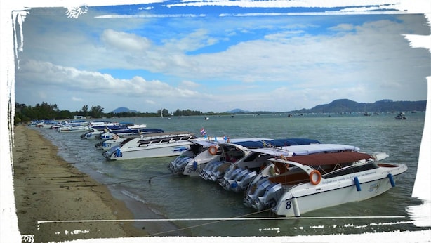 Vários barcos atracados ao longo de uma praia arenosa, com mar calmo e montanhas ao longe.