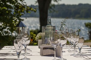 Mesa posta para refeição com copos de vinho, uma garrafa e talheres, com vista para um corpo d'água.