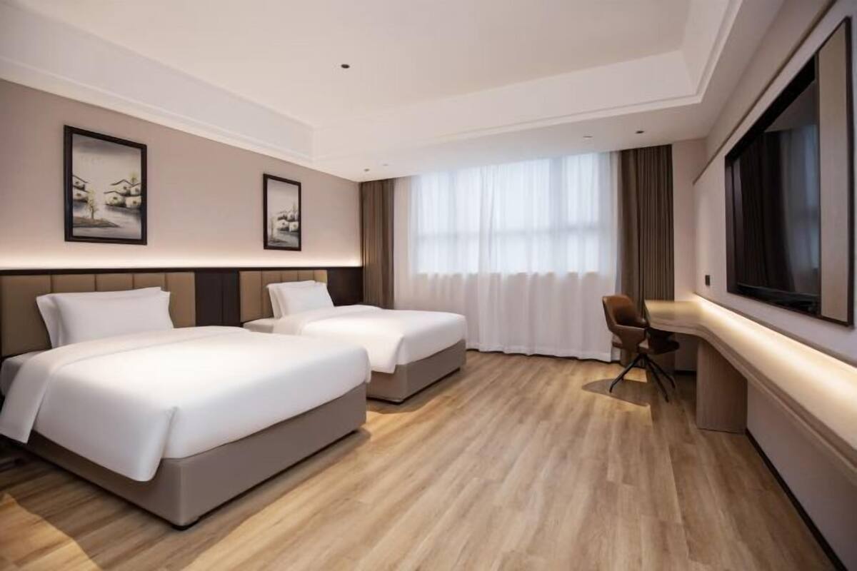 Quarto de hotel com duas camas, uma escrivaninha, uma cadeira, uma janela grande com cortinas e obras de arte nas paredes.