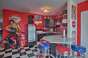 Uma cozinha com tema retrô, com piso xadrez, armários vermelhos e pretos, uma geladeira Coca-Cola e uma estátua de jukebox.