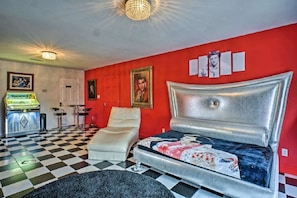 Um quarto com chão xadrez, uma cama com cabeceira metálica, uma parede vermelha e um jukebox.