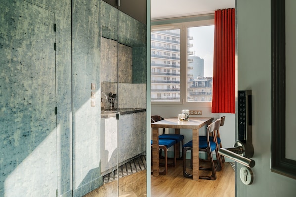 Cozinha moderna com fogão embutido, área de jantar com mesa de madeira e cadeiras azuis, e vista para a cidade através de uma janela com cortinas vermelhas.
