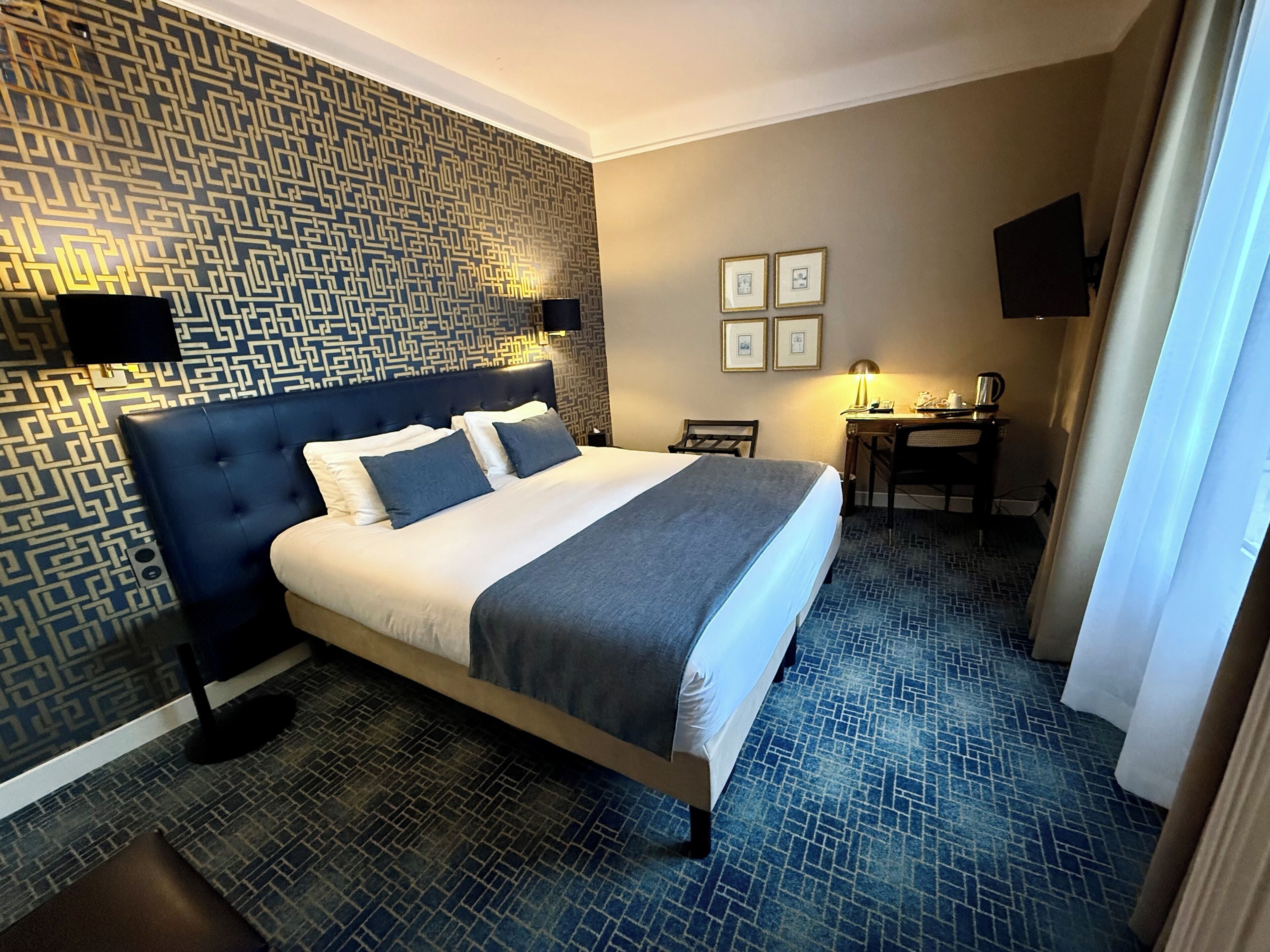 Quarto de hotel com uma cama grande, uma escrivaninha, uma cadeira, uma televisão e parede estampada.