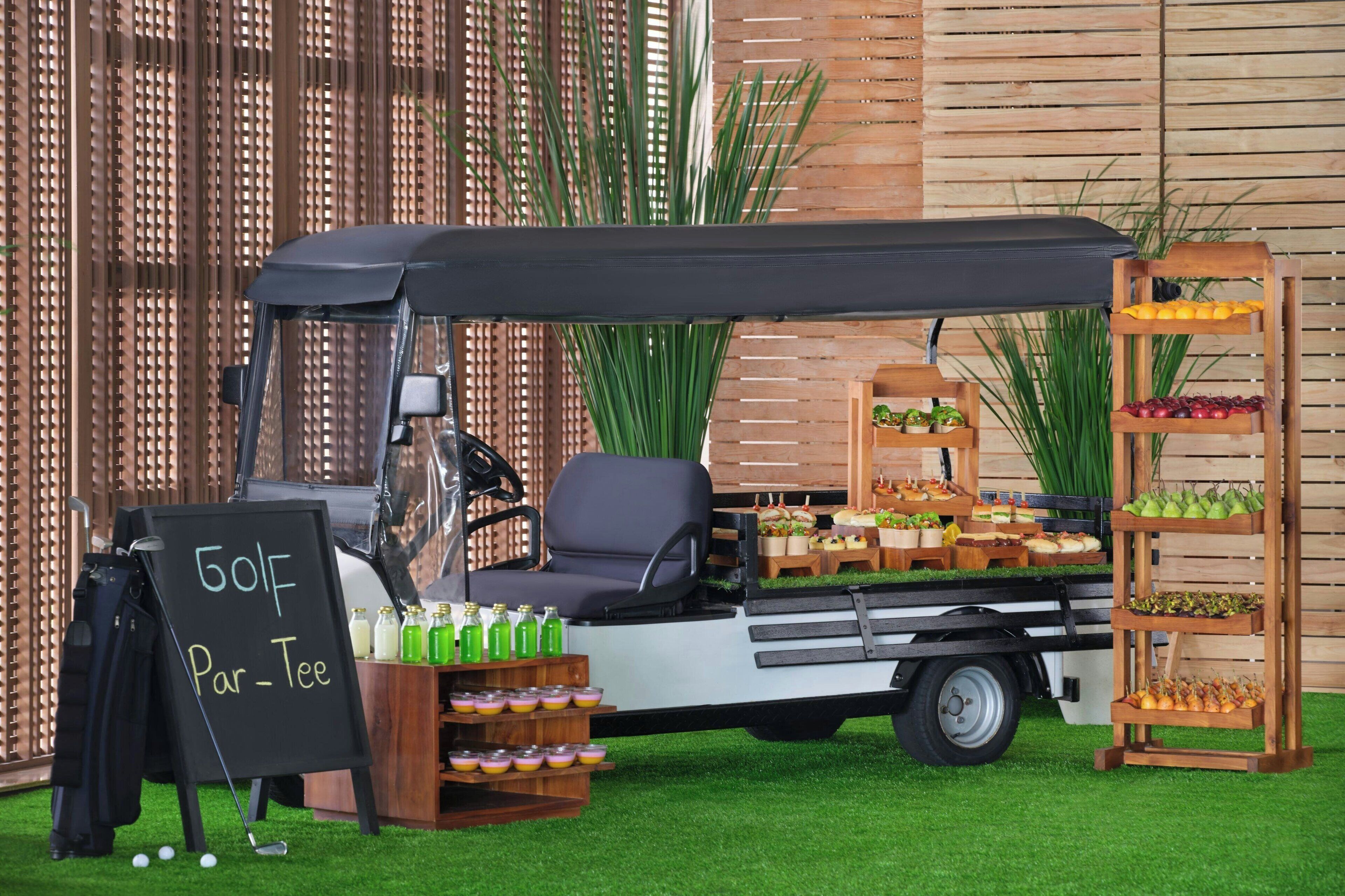 Carrinho de golfe com toldo preto, placa indicando "Golf Par-Tee" e exposição de comida e bebidas.